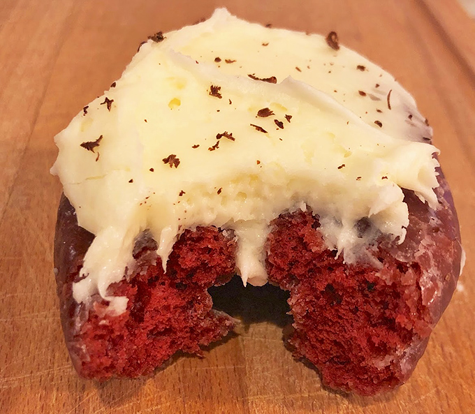A red velvet donut with cream cheese frosting, dramatically halved to reveal its ruby interior. Like cutting into culinary jewelry, with tastier results.