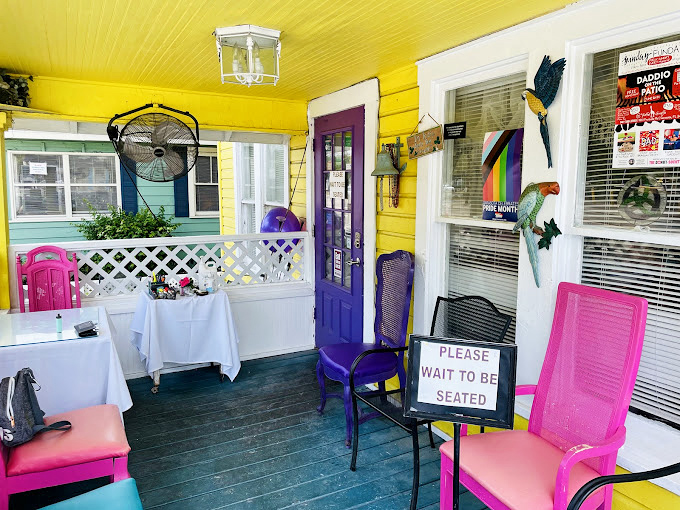 The porch waiting area feels like your fun aunt's Florida home—bright colors, comfortable seating, and a "please wait to be seated" sign that's more invitation than instruction.