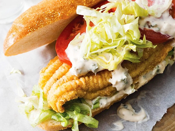 Bisected on its wooden platter, this fish sandwich reveals its generous proportions—flaky white fish nestled with crisp lettuce in a soft bun embrace.