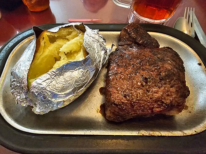 A perfectly seared steak beside a foil-wrapped potato bursting with fluffy goodness. This isn't just dinner&mdash;it's a celebration on a metal tray.