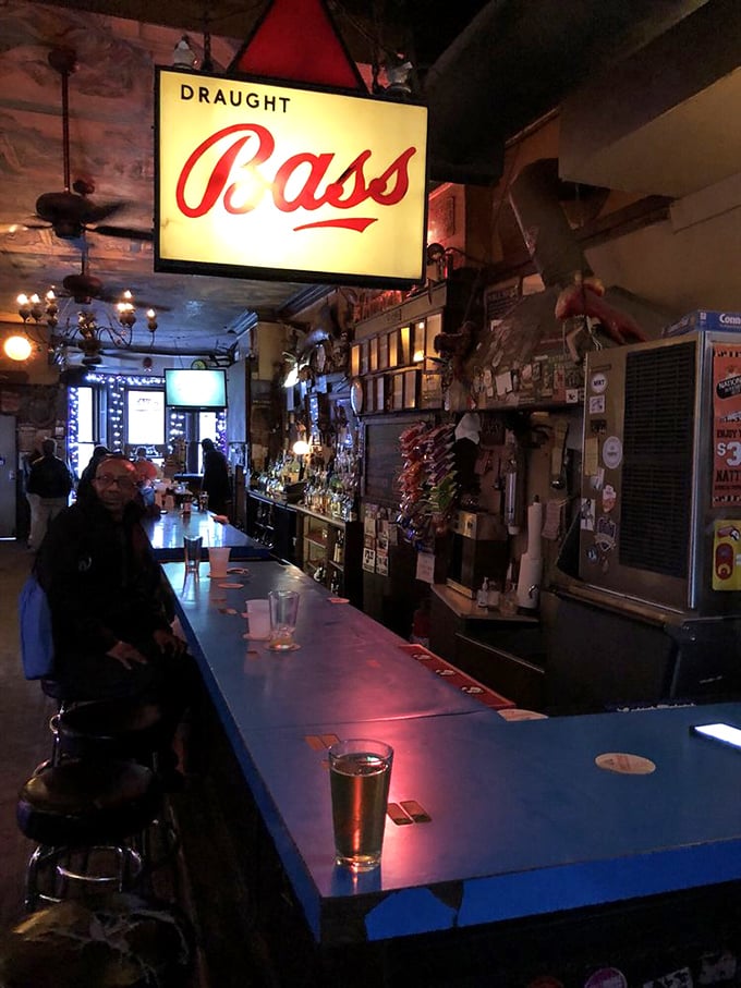 Under the warm glow of a Bass sign, the blue bar counter has supported elbows, drinks, and life stories for decades.