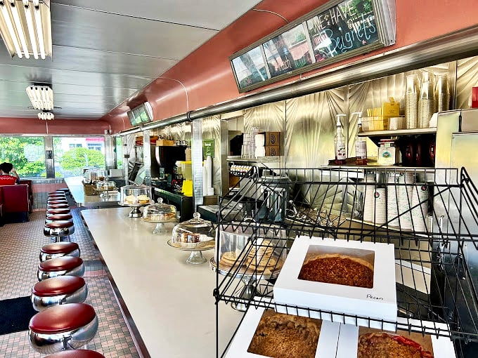 The counter stretches like a runway for comfort food, where pie domes protect their precious cargo and every stool promises a front-row seat to culinary theater.