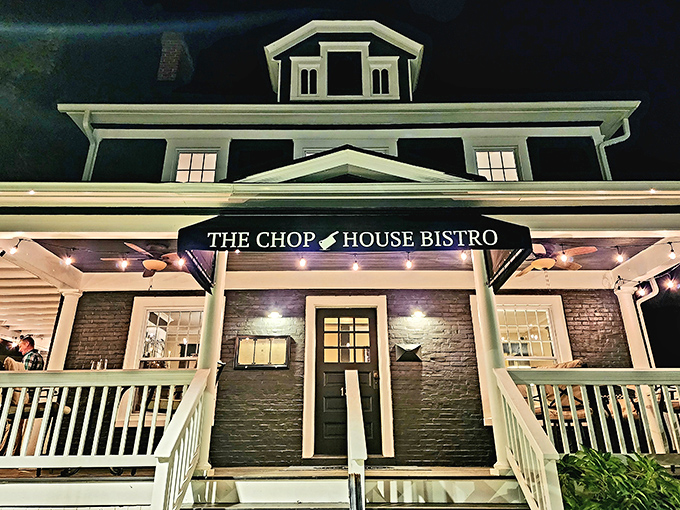 As night falls, Chop House Bistro transforms into a glowing beacon of culinary promise, its white trim and porch lights inviting you in for dinner magic.