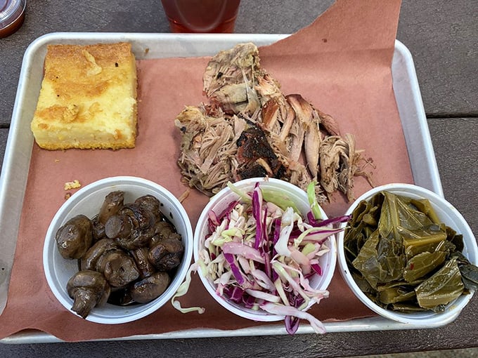 A metal tray of pulled pork perfection flanked by the holy trinity of barbecue sides: mushrooms, slaw, and collards. That cornbread isn't just a side&mdash;it's an essential tool.