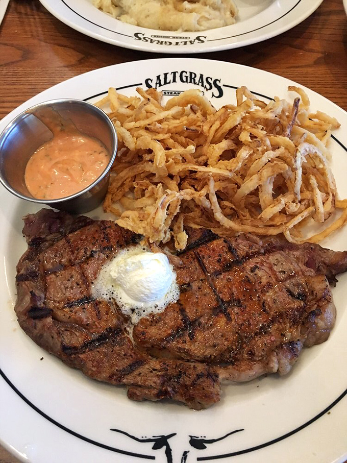 A perfectly grilled steak topped with butter, accompanied by crispy onion strings and dipping sauce—proof that sometimes the simplest pleasures are the most profound.