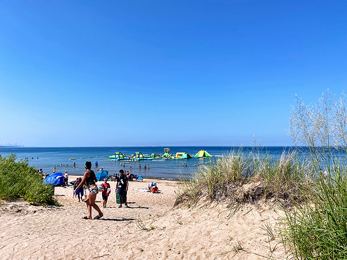 Sandy pathways lead to aquatic thrills. Beach grasses frame this perfect summer tableau where families make their way toward the colorful floating playground offshore.