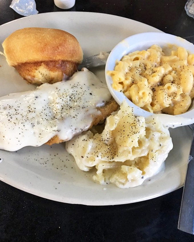 Comfort on a plate: creamy mashed potatoes, buttery roll, and mac and cheese flanking what appears to be country-fried perfection under a blanket of gravy.
