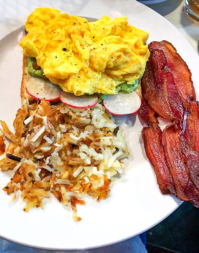 Breakfast elevated: Fluffy scrambled eggs atop avocado toast with crispy hash browns and bacon that would make any morning person out of you.