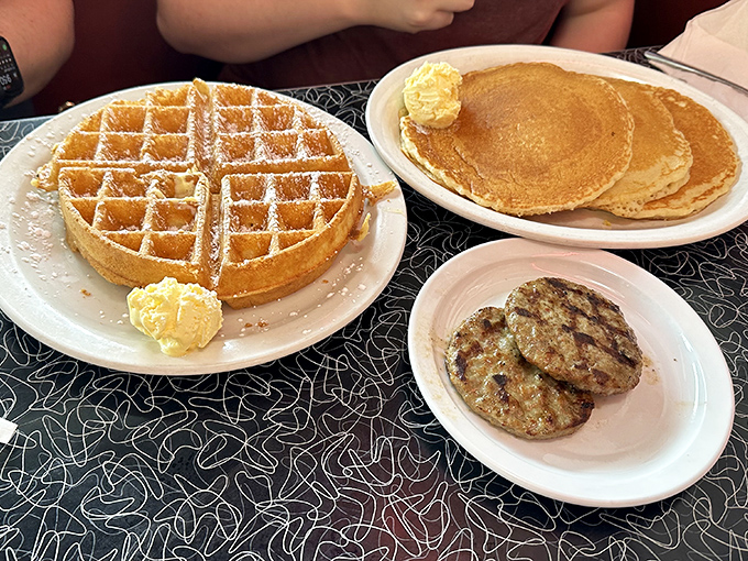The waffle's perfect grid holds pools of melting butter like tiny flavor reservoirs, while those pancakes stand ready for their maple baptism.