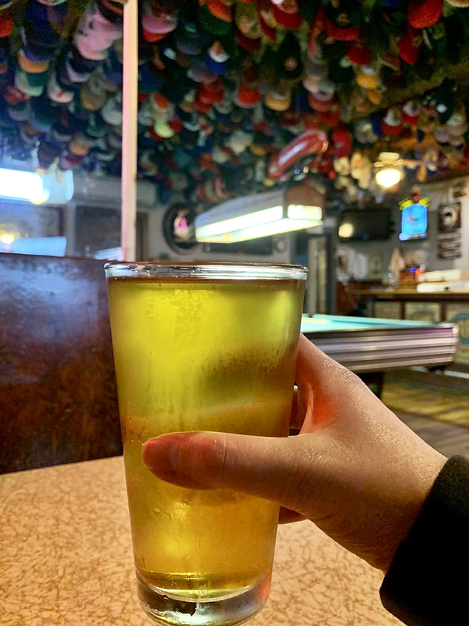 A cold beer catching light beneath the cap collection—liquid gold that's traveled mere miles to meet its burger soulmate.