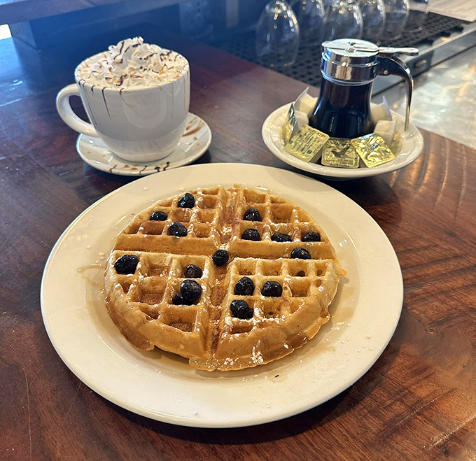 This waffle isn't just breakfast, it's architecture—a perfect grid of golden squares supporting blueberries and maple syrup. The whipped cream coffee is just showing off.