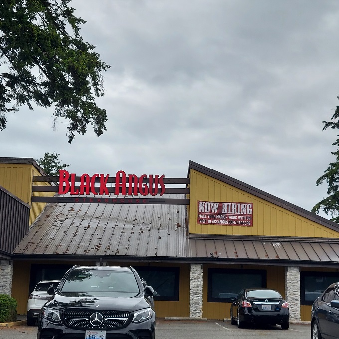 The glowing red signage of Black Angus beckons hungry travelers like a carnivorous lighthouse guiding ships to beefy salvation.