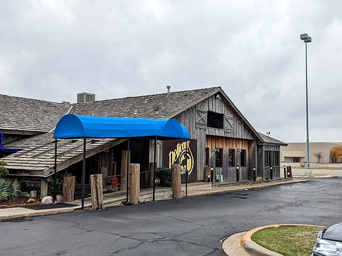 The weathered wooden exterior of Pelican's Restaurant stands like a coastal mirage in Midwest City, complete with signature blue awning and rustic charm.