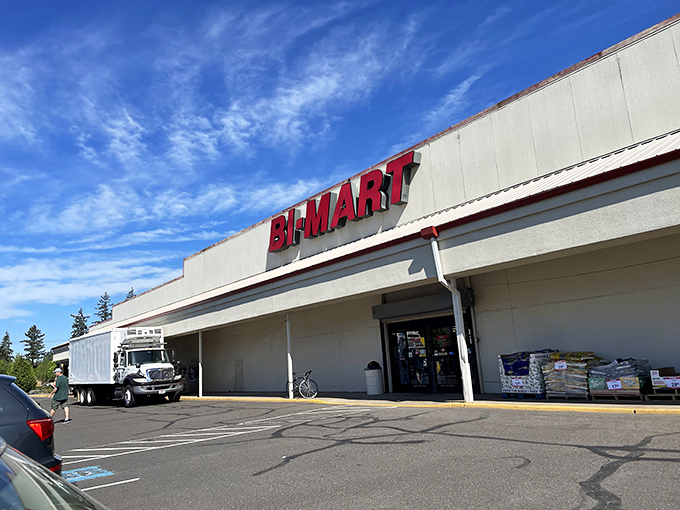 The iconic red Bi-Mart sign against a blue Oregon sky promises treasure hunting adventures within. Garden plants stand ready for your next backyard project.