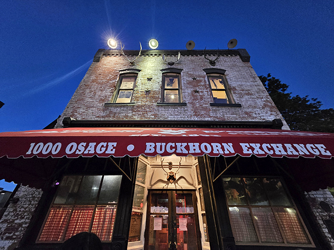 The brick facade of Buckhorn Exchange glows under night lights, its iconic red awning beckoning hungry travelers like a carnivorous lighthouse in urban Denver.