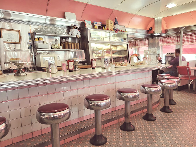 The counter where magic happens&mdash;gleaming stainless steel, cherry-red stools, and the promise of coffee that never reaches the bottom of your cup. 