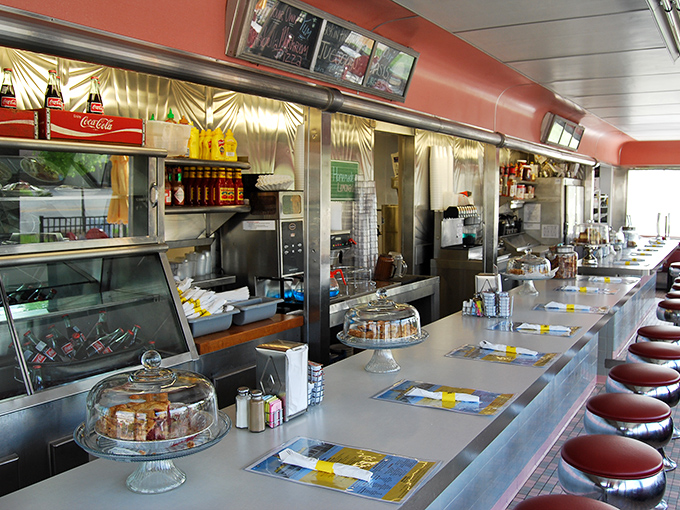 The counter view reveals the heartbeat of diner operations, where pie domes stand sentinel and every seat promises a front-row view of culinary theater.
