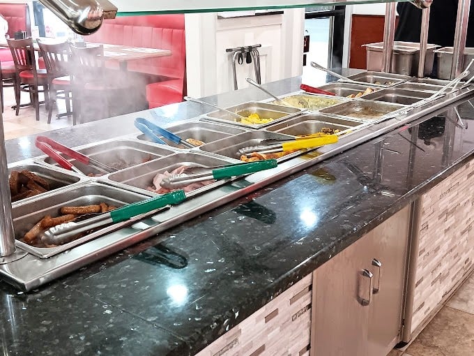 The buffet line&mdash;where decisions become difficult and restraint goes to die. Each metal tray holds promises that your diet made peace with before walking through the door.