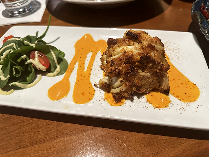 This isn't just a crab cake &ndash; it's a golden-crusted island of seafood perfection swimming in a sunset-colored sauce that demands to be savored.