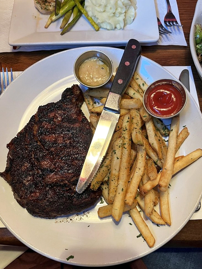 The Delmonico arrives with a crust that crackles with whispered promises, while herb-dusted fries stand ready for their supporting role.