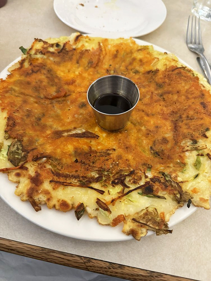 The Korean pancake commands its plate like a crispy-edged monarch, with a small cup of sauce standing guard at the center. 