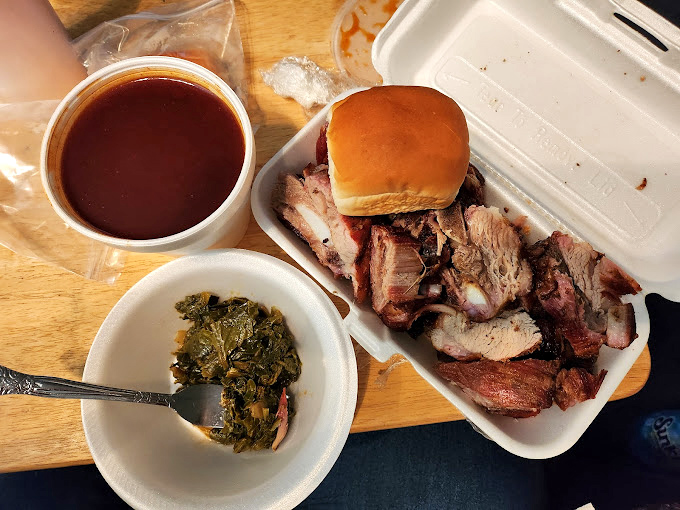 A meat lover's still life: smoke-kissed ribs beside collard greens and sauce. The holy trinity of Southern comfort on a humble styrofoam altar.