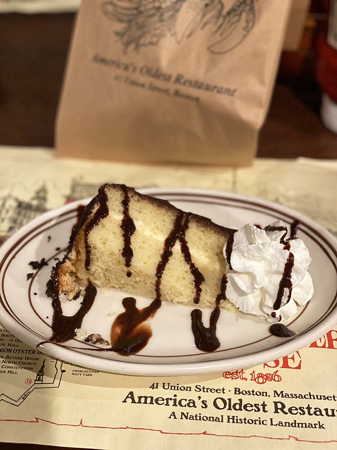 Boston Cream Pie drizzled with chocolate, served on paper proudly proclaiming "America's Oldest Restaurant" &ndash; dessert with a side of heritage.