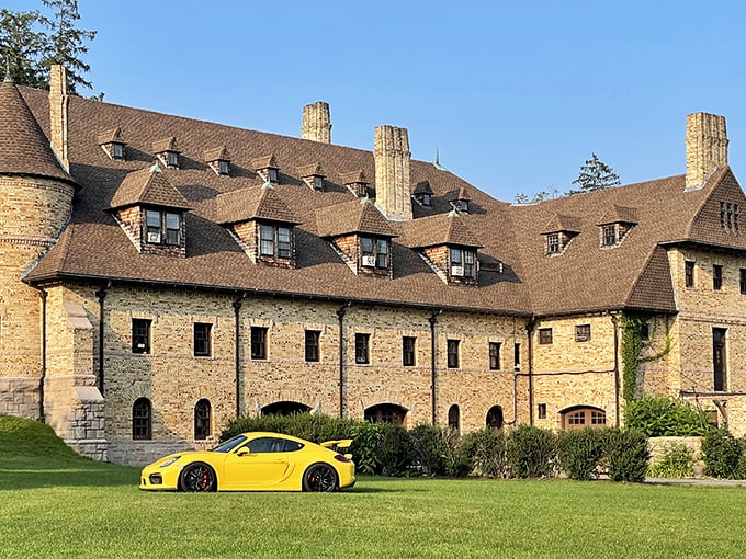 The castle-like exterior of Larz Anderson Auto Museum stands majestically against a blue sky, while a modern yellow Porsche offers a striking contrast to the historic stone facade.