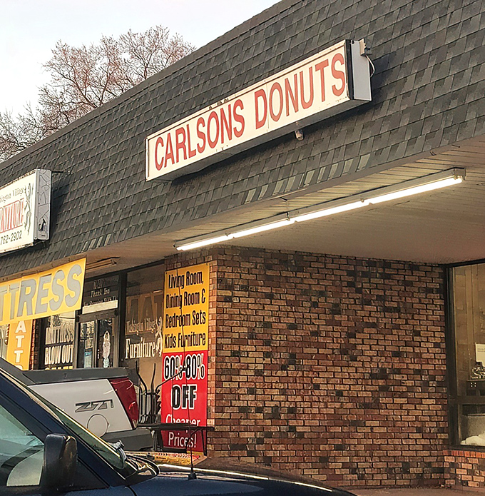 The unassuming strip mall exterior of Carlson's Donuts &ndash; where culinary magic happens behind brick walls and modest signage. Food paradise rarely announces itself with neon.