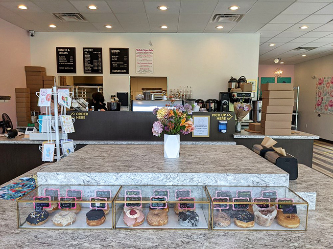 The display case altar where donut dreams materialize, with fresh flowers because these pastries deserve nothing less.