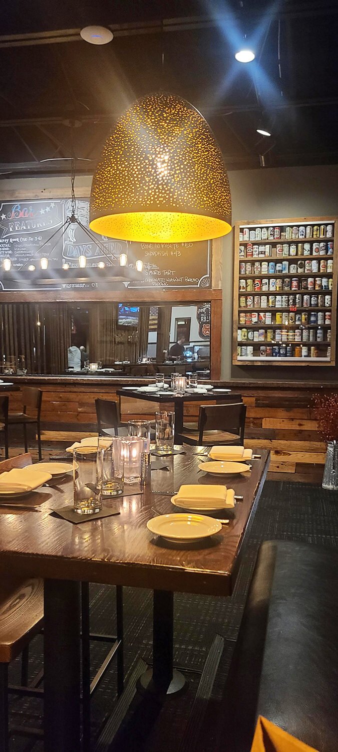 A golden perforated pendant lamp casts honeycomb shadows across the dining area while a wall of spirits promises liquid courage before your carnivorous adventure.