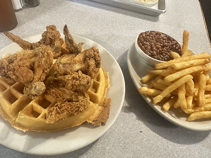 The main event arrives: crispy, golden-brown chicken perched atop a fluffy waffle, with French fries and beans standing by.