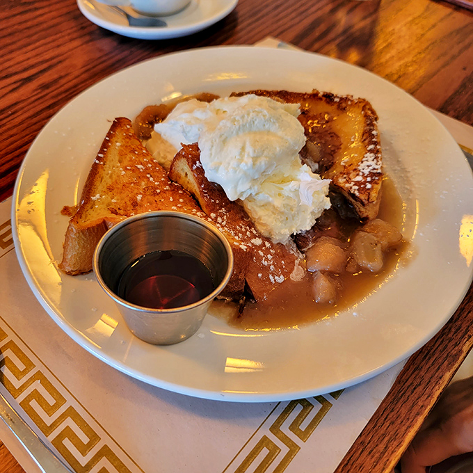Breakfast architecture at its finest: golden-brown French toast supporting a cloud of whipped cream, with maple syrup waiting in the wings.