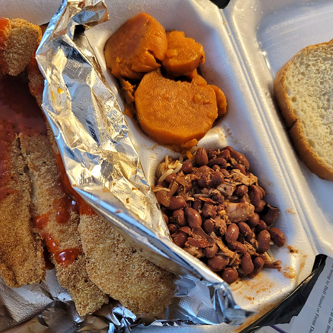 Sweet candied yams and perfectly seasoned beans share space with cornmeal-crusted catfish. A holy trinity of Southern comfort on a humble styrofoam throne.