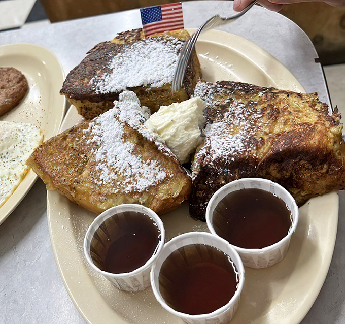 This French toast extravaganza arrives like breakfast royalty&mdash;powdered sugar snowfall, whipped cream cloud, and three cups of syrup because some decisions should be personal.