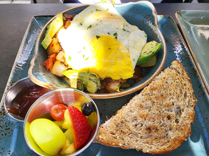 A sunny-side egg grins atop a mountain of hash, with fruit standing by like a nutritional conscience whispering, "You're doing great!"