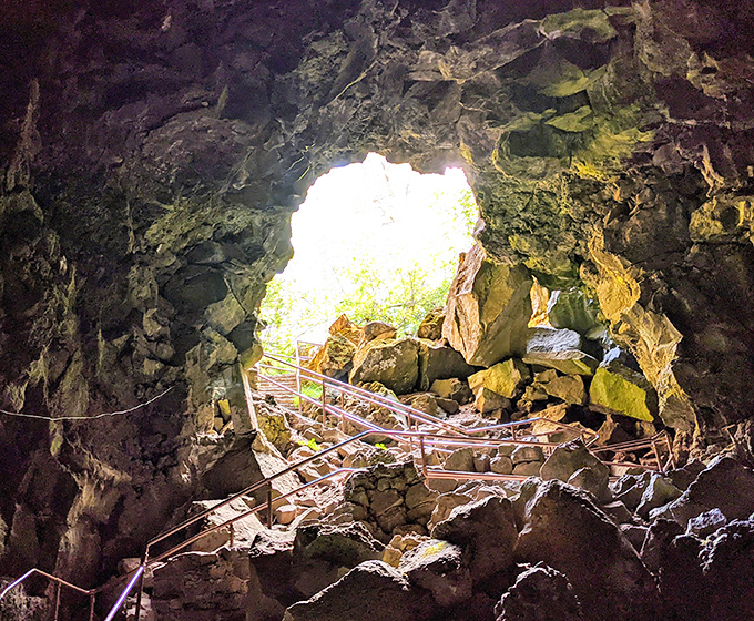 The dramatic entrance to Lava River Cave feels like stepping into another world. Sunlight streams through the opening, illuminating ancient volcanic rock formations and metal stairs descending into darkness.