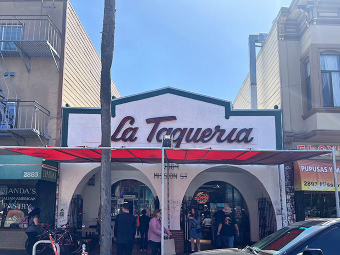 The iconic white facade with that bold red script is like a beacon for burrito pilgrims. La Taqueria's exterior promises culinary magic within those arched entryways.
