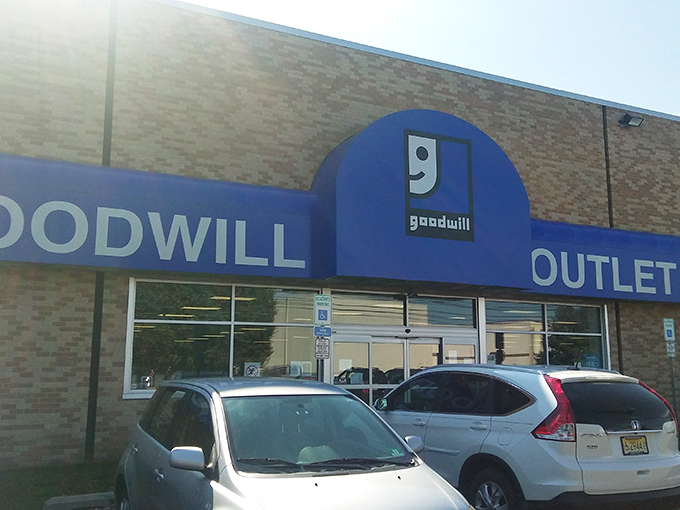 The blue Goodwill sign beckons like a lighthouse to bargain hunters navigating the seas of retail markup. Behind these doors, treasure awaits.