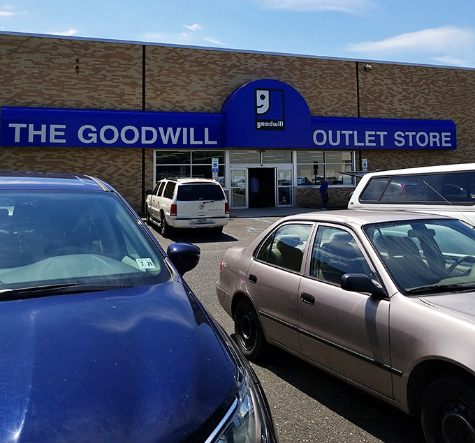 The blue Goodwill sign beckons like a lighthouse to bargain hunters navigating the seas of retail markup. Behind these doors, treasure awaits.