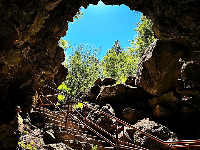 The dramatic entrance to Lava River Cave feels like stepping into another world. Sunlight streams through the opening, illuminating ancient volcanic rock formations and metal stairs descending into darkness.