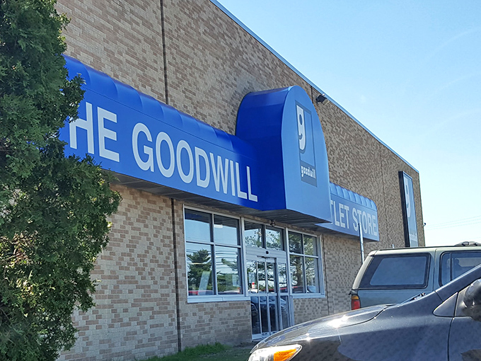 The blue Goodwill sign beckons like a lighthouse to bargain hunters navigating the seas of retail markup. Behind these doors, treasure awaits.