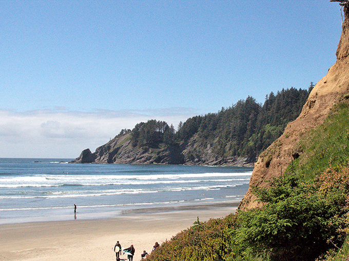 Nature's perfect postcard – dramatic headlands embrace golden sands while surfers dance on gentle waves beneath an impossibly blue Oregon sky.