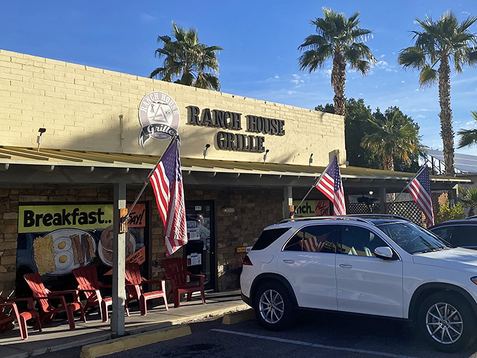 The unassuming exterior of Ranch House Grille, where American flags flutter in welcome and palm trees stand guard over breakfast paradise.