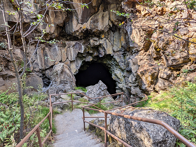 The dramatic entrance to Lava River Cave feels like stepping into another world. Sunlight streams through the opening, illuminating ancient volcanic rock formations and metal stairs descending into darkness.