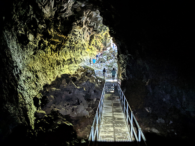 The dramatic entrance to Lava River Cave feels like stepping into another world. Sunlight streams through the opening, illuminating ancient volcanic rock formations and metal stairs descending into darkness.