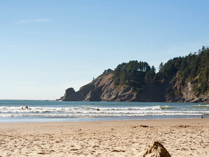 Nature's perfect postcard &ndash; dramatic headlands embrace golden sands while surfers dance on gentle waves beneath an impossibly blue Oregon sky.