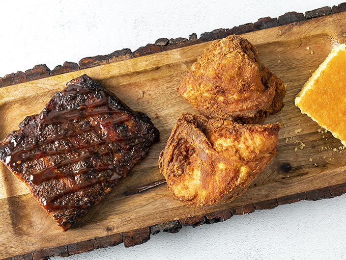 A wooden board showcasing the holy trinity of Southern indulgence: sticky-sweet ribs, golden-crusted chicken, and cornbread that tastes like sunshine.