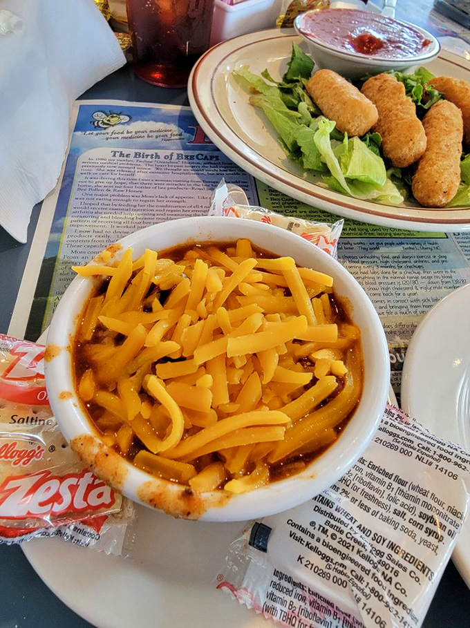 A bowl of chili topped with enough cheese to make Wisconsin proud, sitting beside what appears to be mozzarella sticks on a bed of lettuce.