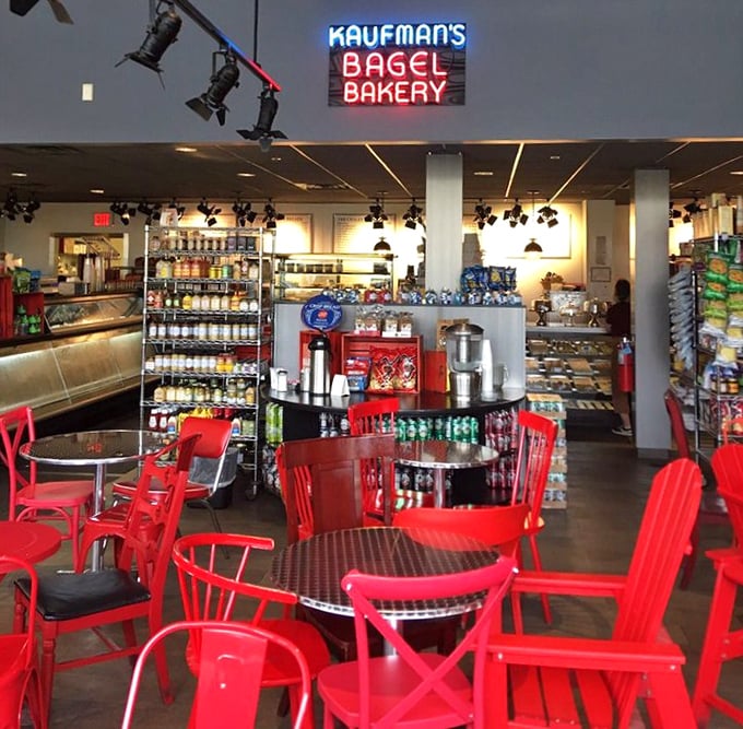 Red chairs pop against the monochromatic backdrop, creating a casual dining space. The neon sign glows like a beacon for bagel pilgrims.